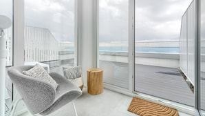 Uma varanda moderna com vista para o mar, uma cadeira branca com almofadas e um banquinho de madeira.