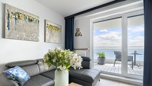 Uma sala de estar moderna com um sofá de couro cinza, uma mesa de centro de madeira e um vaso com flores. Há duas pinturas abstratas na parede, uma cortina azul e vista para o mar através de grandes portas de correr.