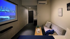 Quarto de hotel com uma tela grande exibindo uma paisagem urbana, um sofá, uma mesa pequena com uma planta e uma cama.