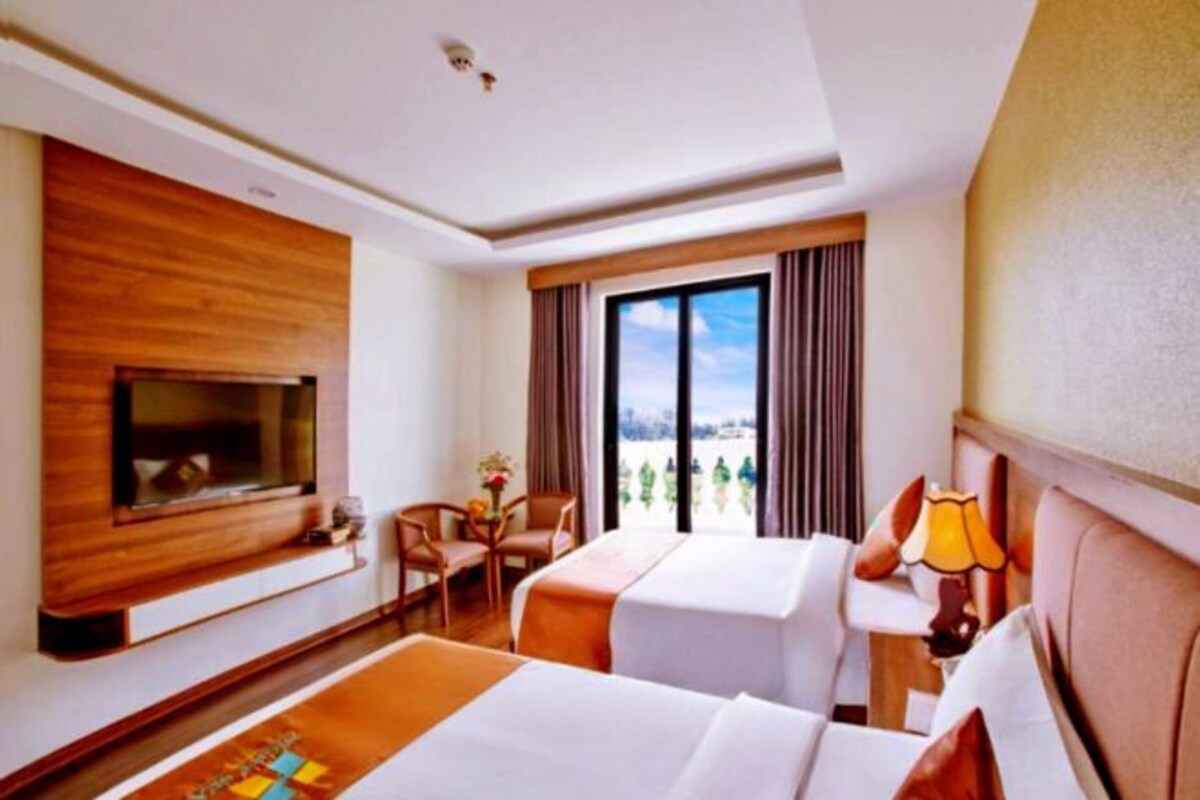 Quarto de hotel com duas camas, uma televisão presa à parede, uma mesa de trabalho com luminária e vista para o exterior.