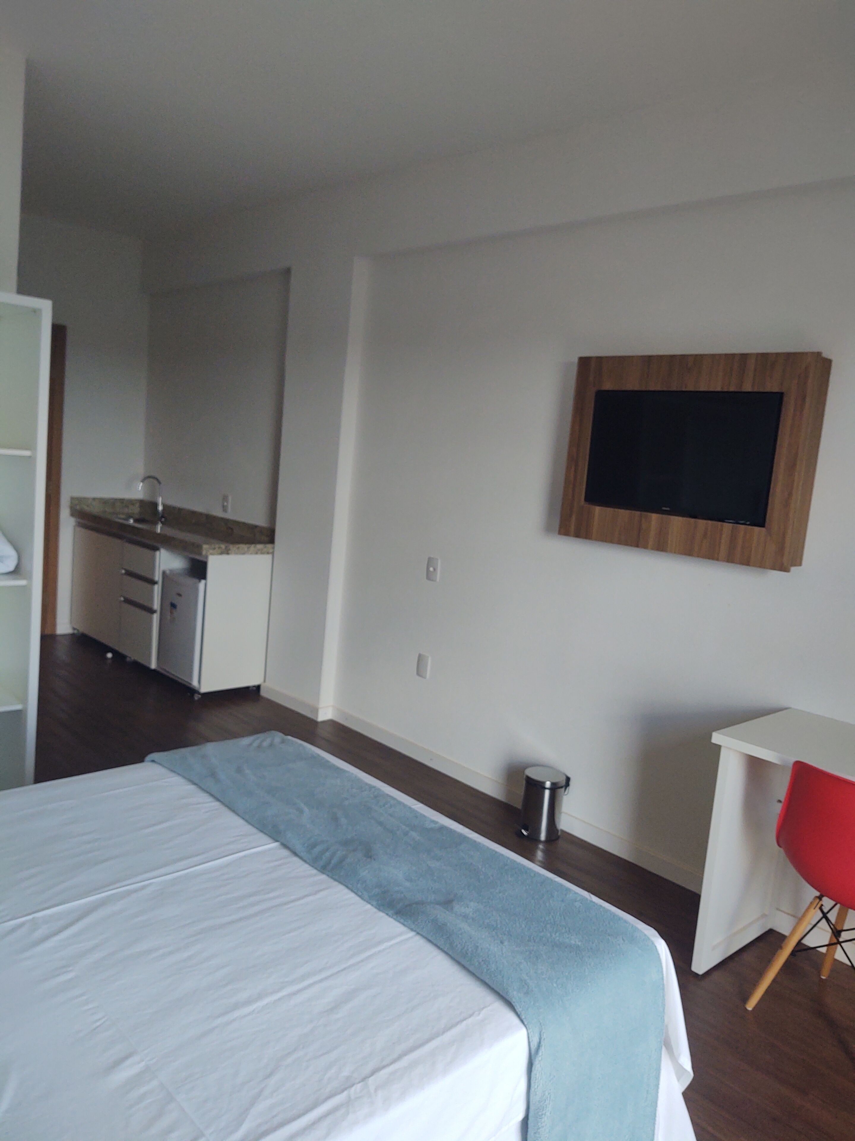 Quarto com cama, escrivaninha e televisão fixada na parede.