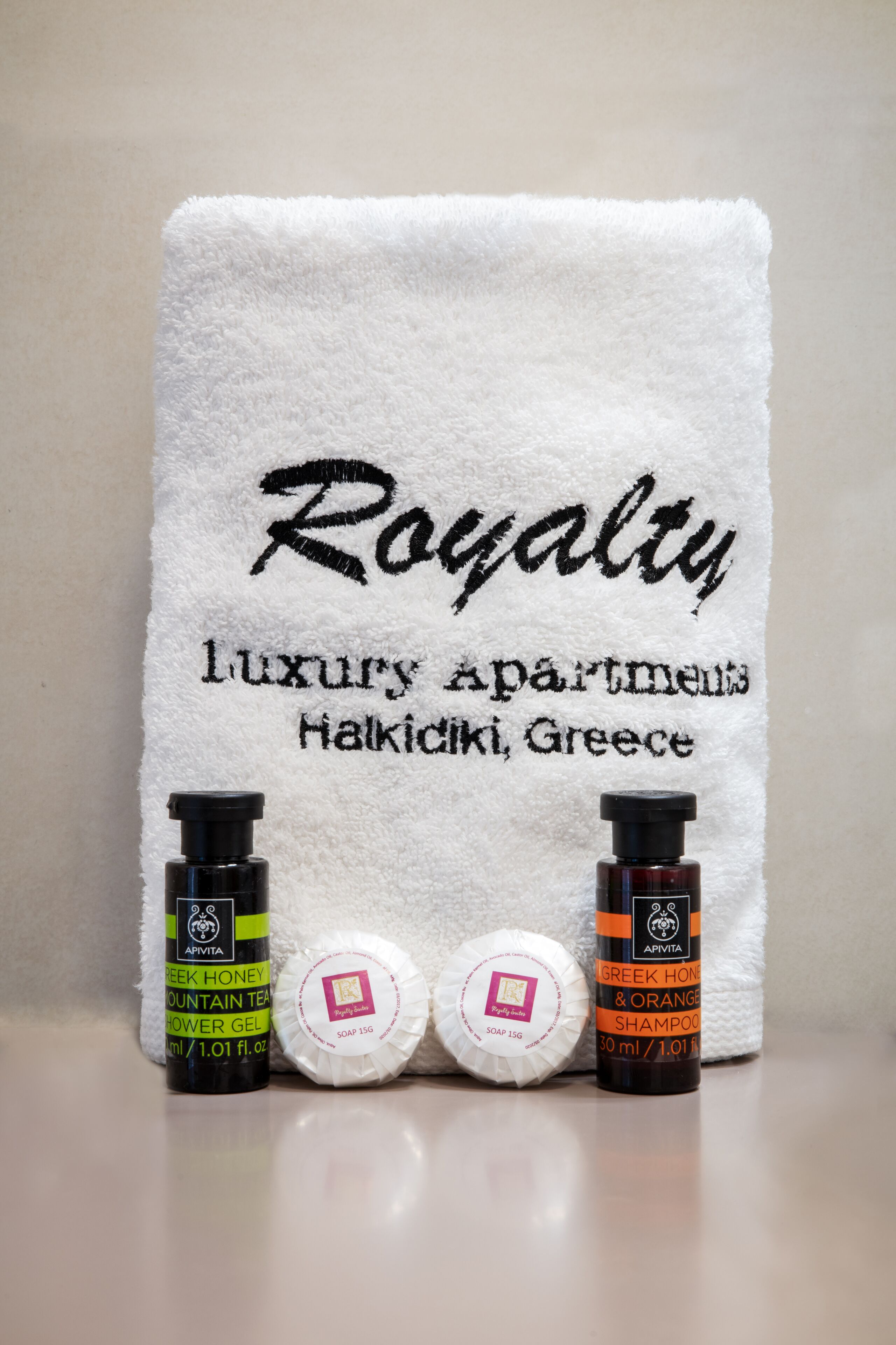 Toalha branca com a inscrição "Royalty Luxury Apartments Halkidiki, Greece", acompanhada de duas garrafas de "Greek Honey & Orange Shampoo" e dois sabonetes.