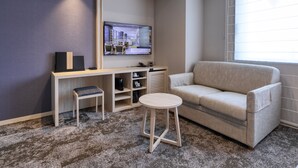 Uma sala de estar moderna com um sofá, uma mesa redonda pequena, uma TV fixada na parede e uma escrivaninha com uma cadeira.