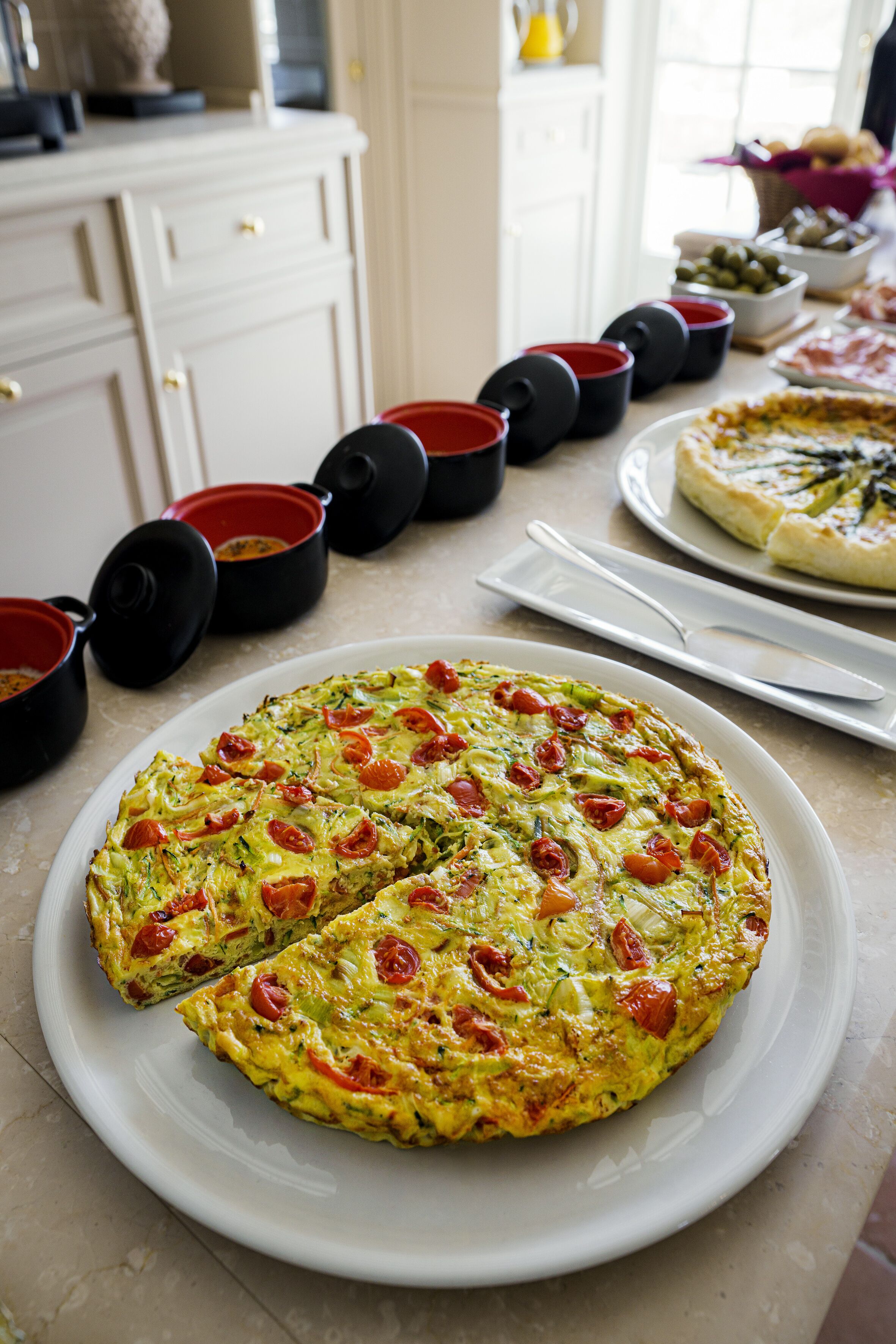 Uma bancada de cozinha com uma quiche grande, várias pequenas tigelas pretas e uma pizza num prato branco.