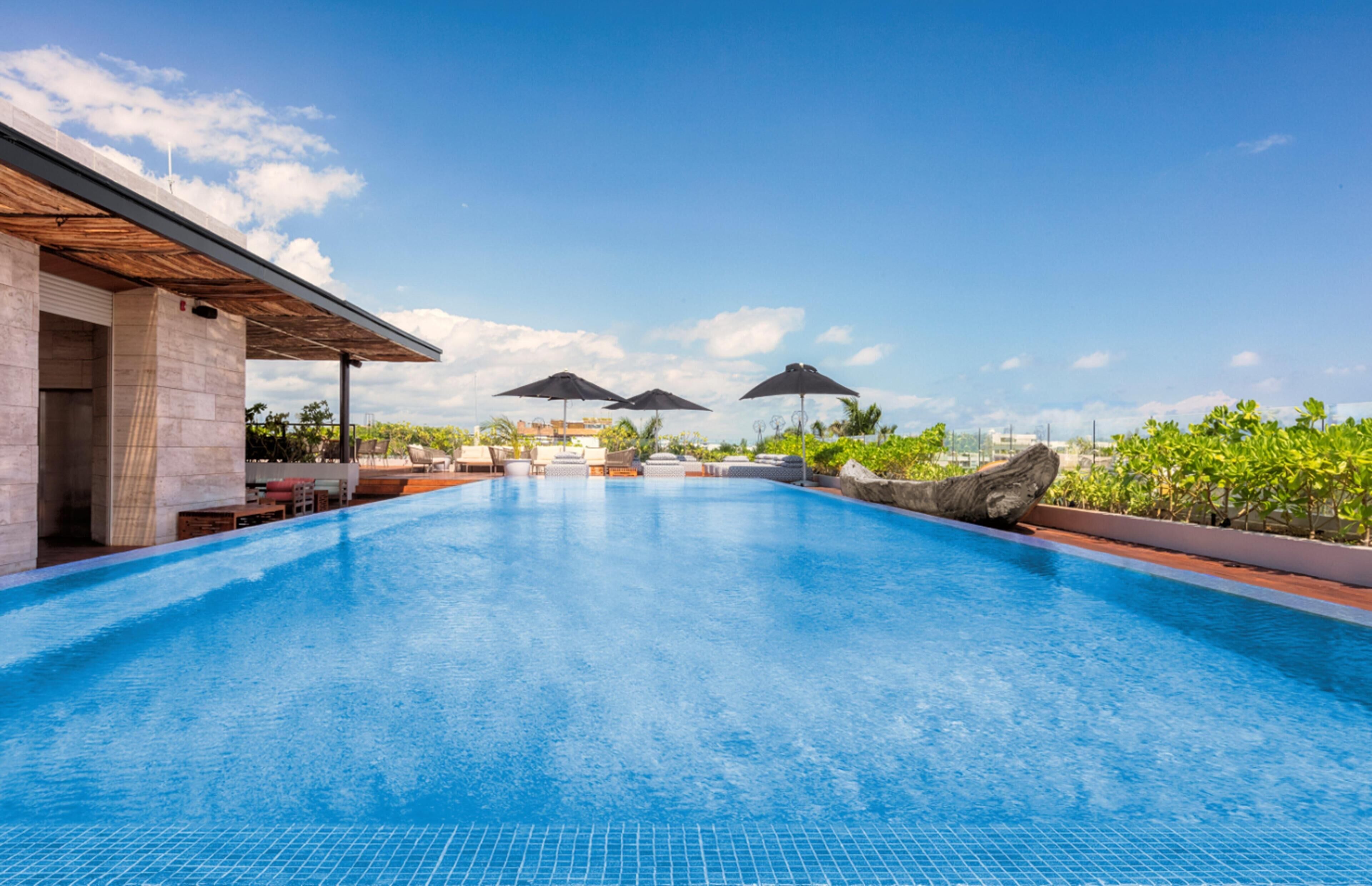 Piscina com superfície de água azul cristalina, rodeada por espreguiçadeiras e guarda-sóis, e um edifício de pedra com área de varanda coberta.