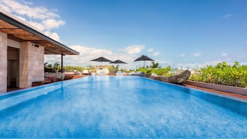 Piscina com superfície de água azul cristalina, rodeada por espreguiçadeiras e guarda-sóis, e um edifício de pedra com área de varanda coberta.