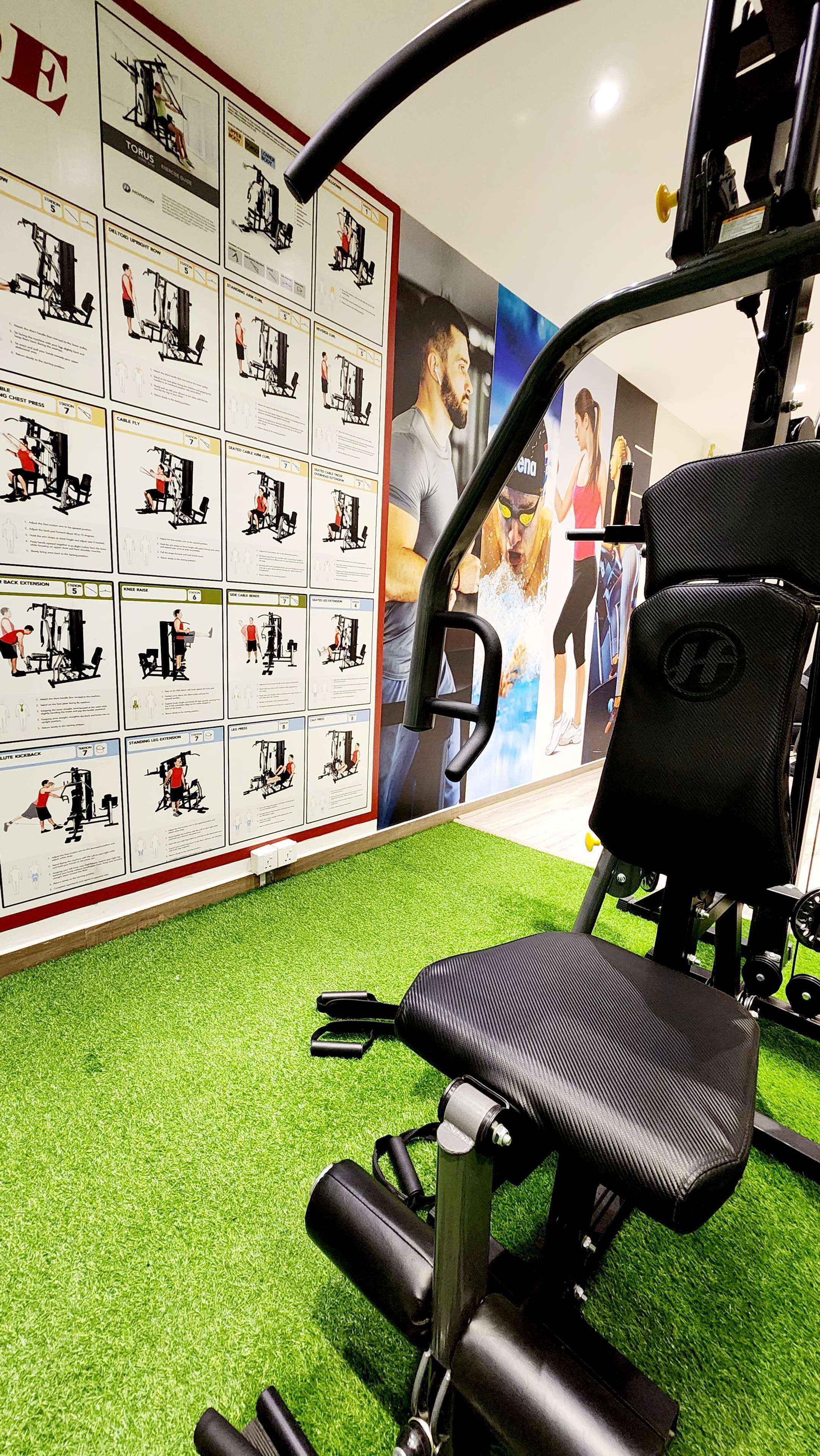 Uma academia com diversos aparelhos de ginástica, uma cadeira preta e uma parede com cartazes de exercícios.