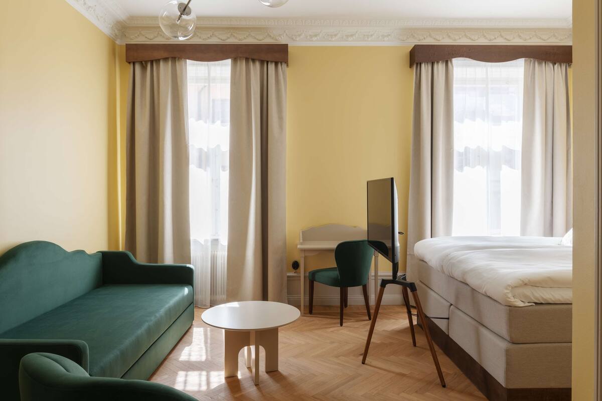 Quarto de hotel com sofá verde, mesa redonda branca, secretária com cadeira e uma cama.