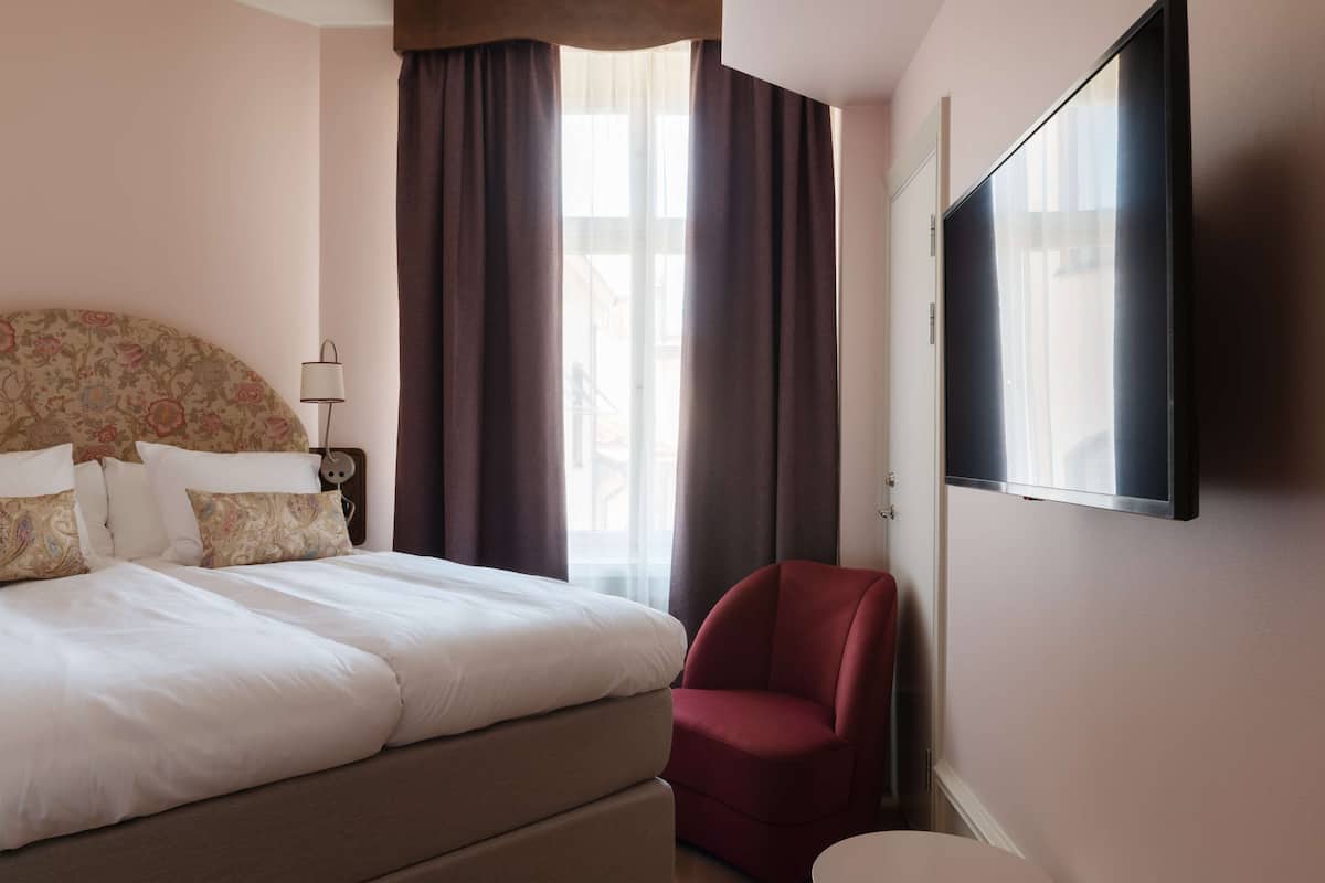 Quarto de hotel com uma cama, uma poltrona vermelha, uma televisão e uma janela com cortinas.