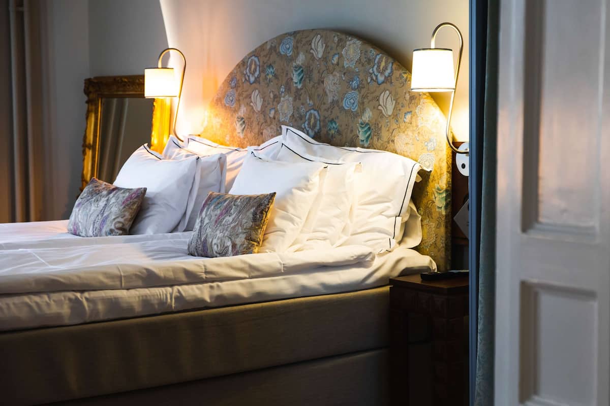 Uma cama bem arrumada com lençóis brancos e almofadas decorativas, uma cabeceira estampada com flores e luminárias de leitura fixadas na parede.