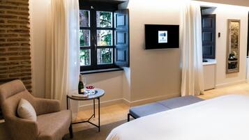 Quarto de hotel com cama, uma cadeira, uma pequena mesa com garrafa e copos, televisão na parede e janela com cortinas.
