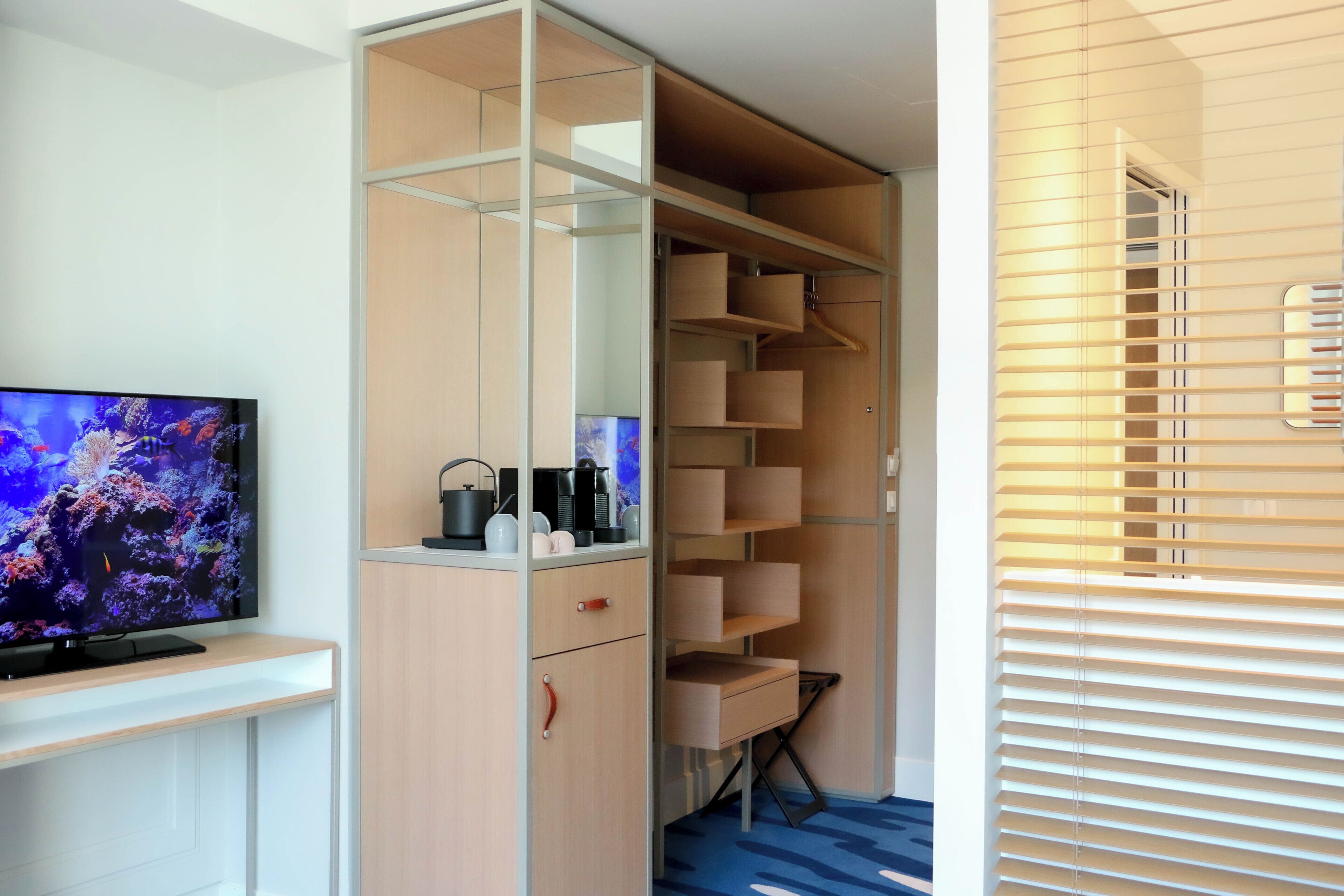 Quarto com televisão, armário de madeira com gavetas, prateleira com objetos e janela com persianas.
