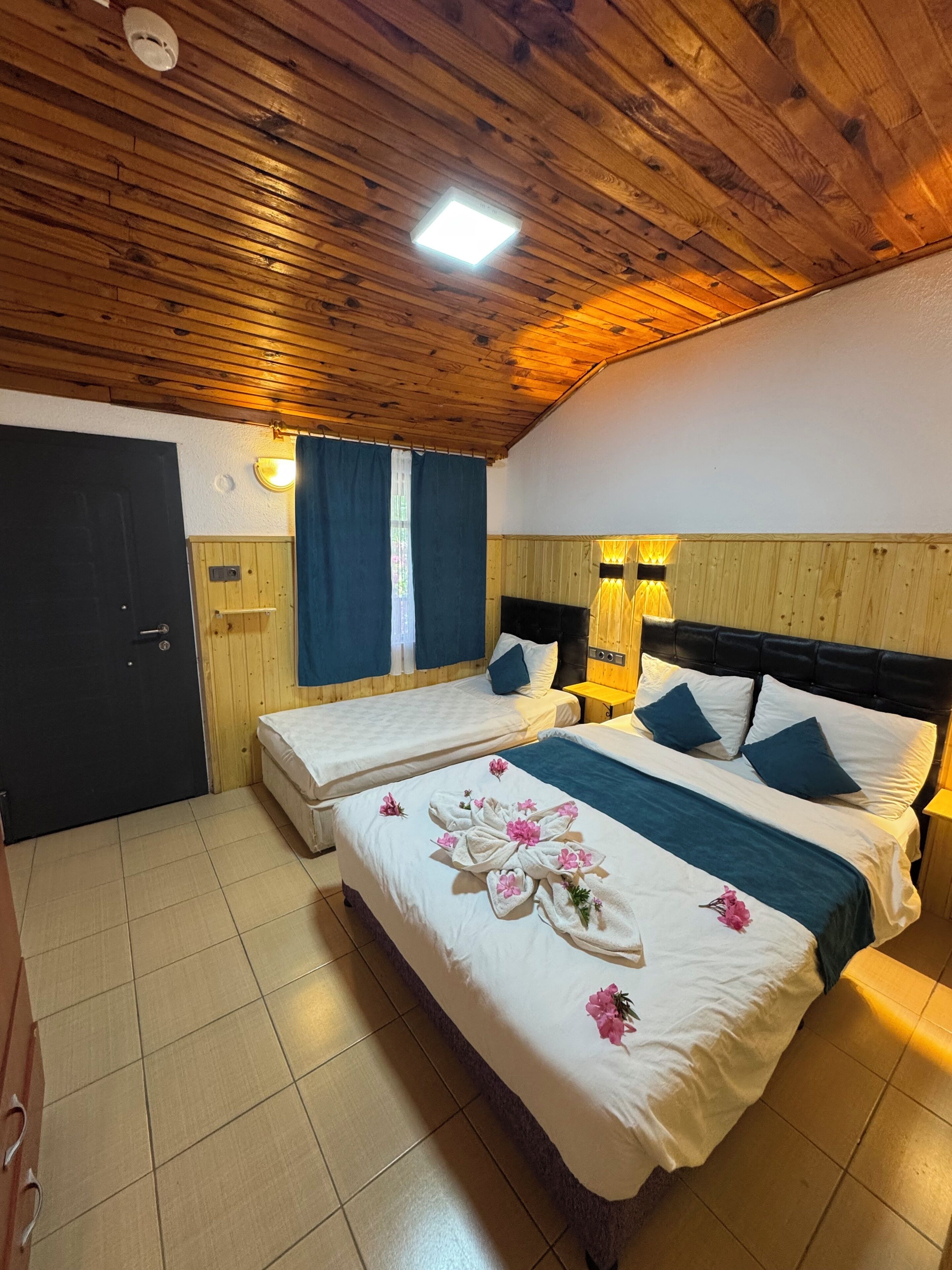 Quarto de hotel com teto de madeira, duas camas, um banheiro pequeno e piso ladrilhado.