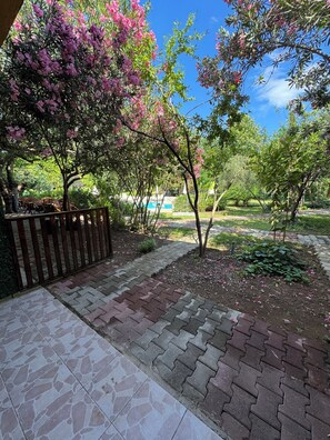 Um caminho pavimentado leva a uma área de estar com cadeiras, rodeada por árvores floridas e uma piscina ao fundo.
