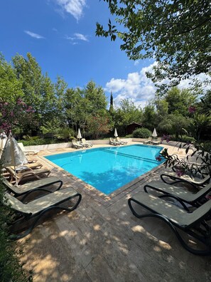 Piscina com espreguiçadeiras e guarda-sóis, cercada por árvores e vegetação.