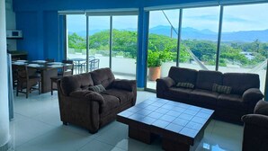 Uma sala de estar moderna com dois sofás, uma mesa de centro e um espaço de jantar com mesa e cadeiras. Grandes janelas oferecem vista para vegetação e montanhas.