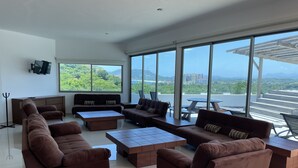 Uma sala de estar moderna com sofás marrons, uma mesa de centro e uma TV. Janelas amplas oferecem vista para áreas verdes e edifícios.