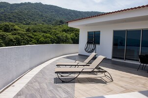 Terraço no telhado com cadeiras de descanso, porta de vidro e vista para uma colina arborizada.