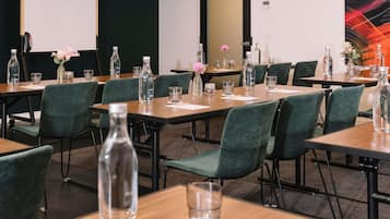 Sala de conferências com mesas, cadeiras, garrafas de água e tela de projeção.