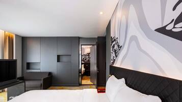 Quarto de hotel moderno com uma cama, escrivaninha, televisão e uma parede com arte abstrata.