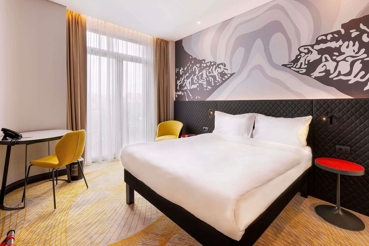 Quarto de hotel com uma cama, uma escrivaninha, duas cadeiras, uma mesinha e um painel decorativo na parede.