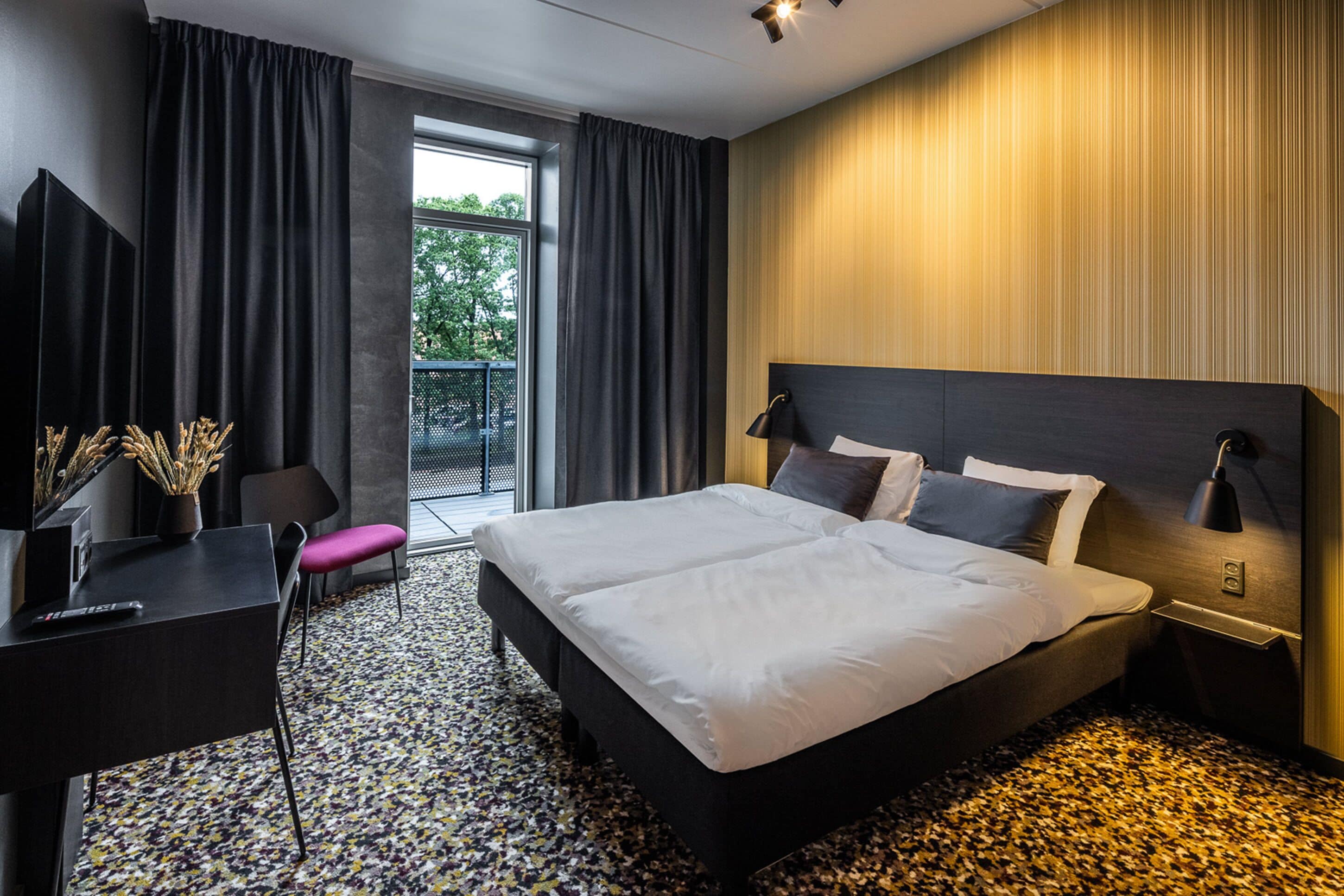 Quarto de hotel moderno com uma cama grande, uma escrivaninha com cadeira, uma televisão e vista para o exterior pela janela.