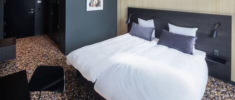 Quarto de hotel com uma cama grande, cabeceira, luminária na parede e piso coberto de carpete.