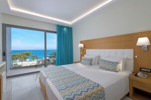 Quarto de hotel moderno com uma cama grande, cabeceira de madeira, mesa de cabeceira com telefone e vista para o mar através de uma porta de vidro deslizante.