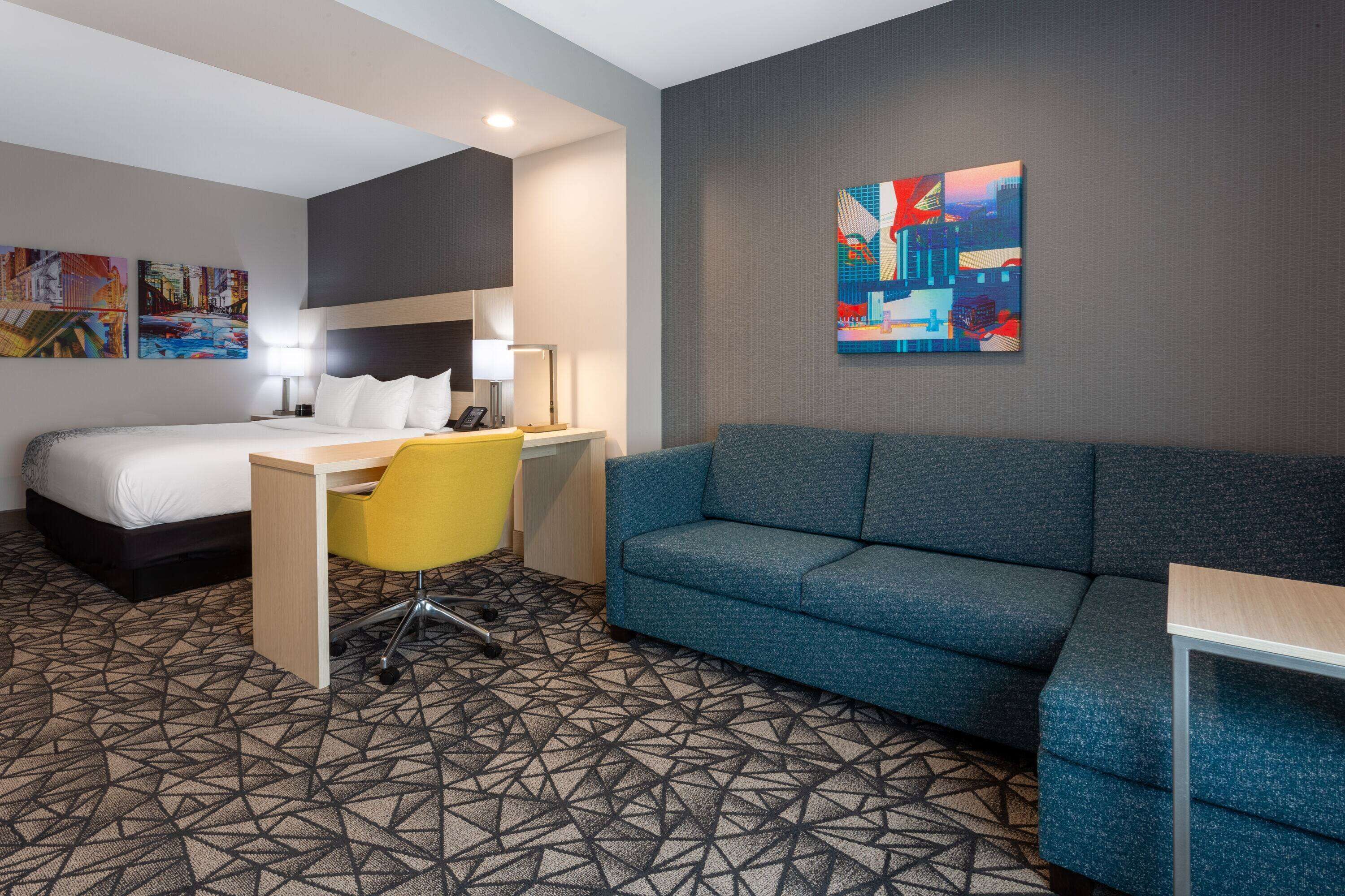 Quarto de hotel com cama, escrivaninha com cadeira, sofá e obras de arte nas paredes.