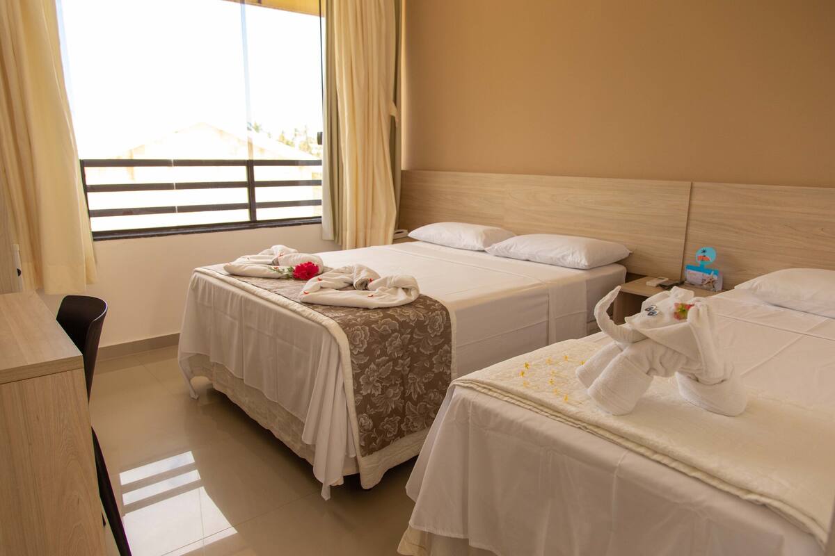 Quarto de hotel com duas camas, uma escrivaninha de madeira e uma janela com cortinas.