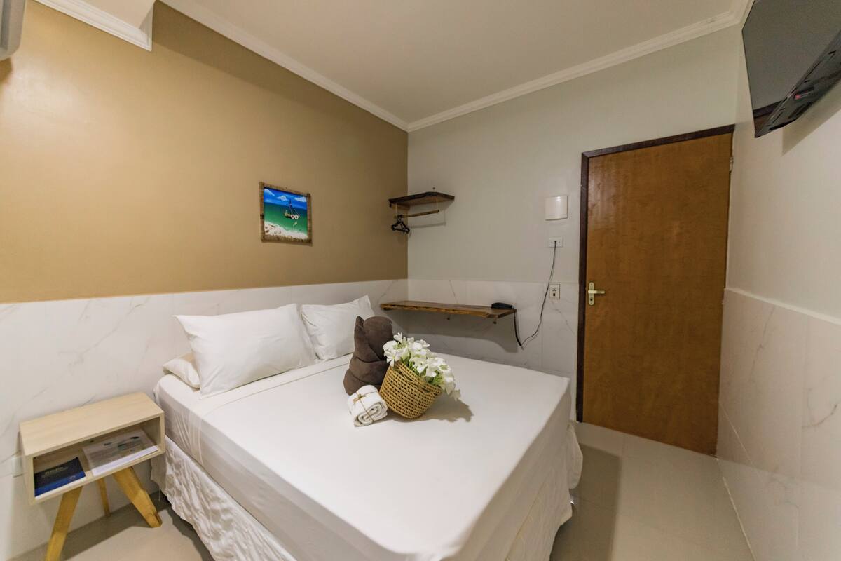 Cama bem arrumada com lençóis brancos, uma mesinha de cabeceira com um livro, uma prateleira na parede com um cesto de flores, e uma porta à direita.