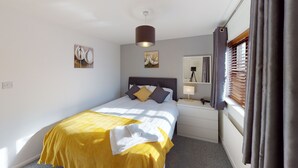 Quarto bem arrumado com cama, mesa de cabeceira, abajur e janela com persianas.