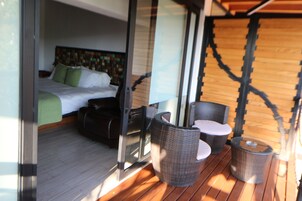 Quarto com cama, almofadas verdes e mala. Há uma cadeira de vime e uma mesa pequena em um deck de madeira.