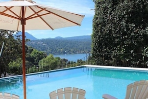 Piscina com espreguiçadeiras e guarda-sol, cercada por vegetação exuberante e vista para um lago e montanhas.