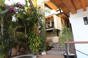 Escadaria que leva a um deck de madeira, com vegetação exuberante e um edifício branco ao fundo.