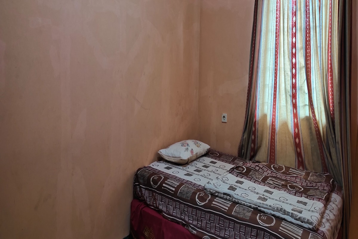 Quarto pequeno com uma cama de solteiro, colcha estampada, travesseiro e cortina.