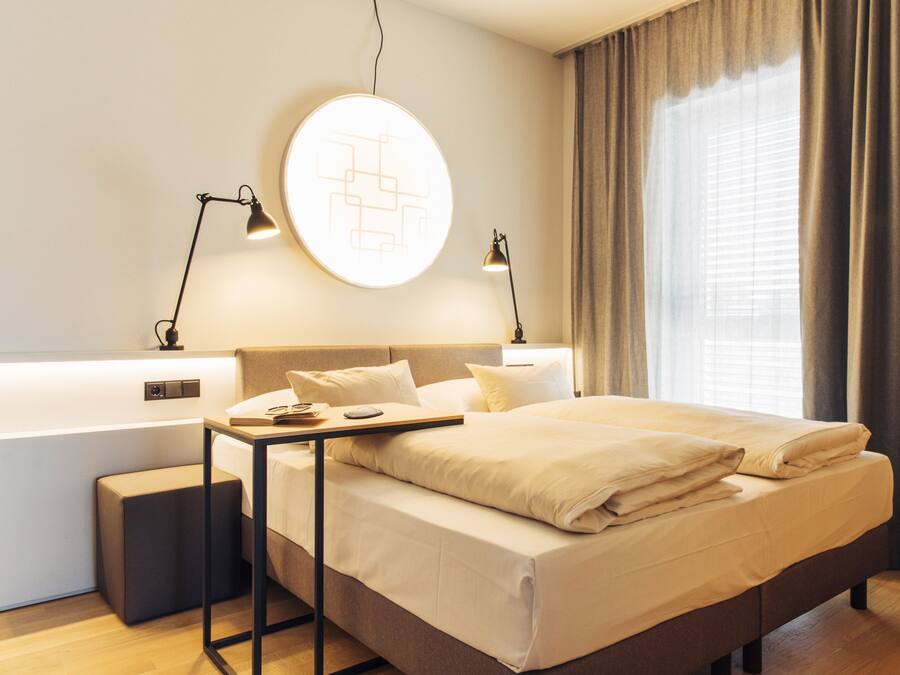 Quarto de hotel moderno com uma cama grande, mesas de cabeceira e um aplique na parede.