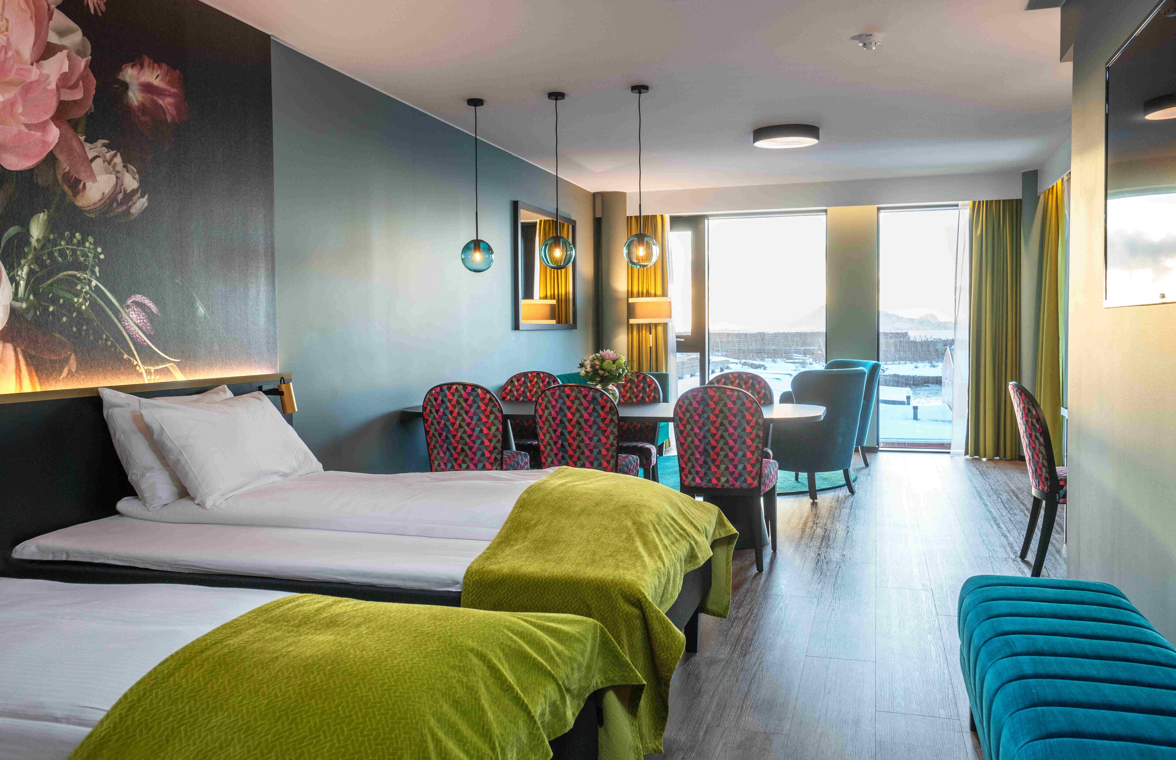 Quarto de hotel com duas camas, uma escrivaninha com cadeiras, uma janela ampla com vista e uma varanda.