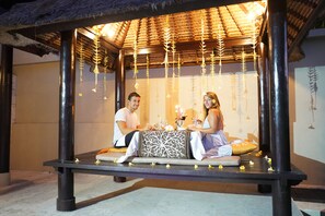 Um casal sentado num banco sob um telhado de palha, rodeado por decorações penduradas e velas.