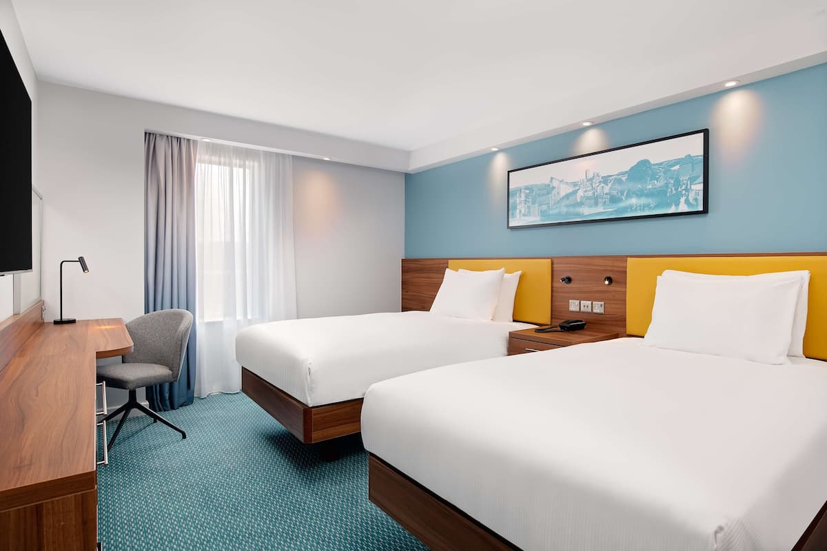 Quarto de hotel com duas camas, uma escrivaninha, uma cadeira e uma janela grande com cortinas.
