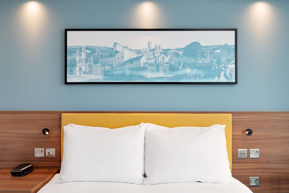 Quarto de hotel com cama, almofadas brancas, fotografia emoldurada de um panorama urbano e cabeceira de madeira.