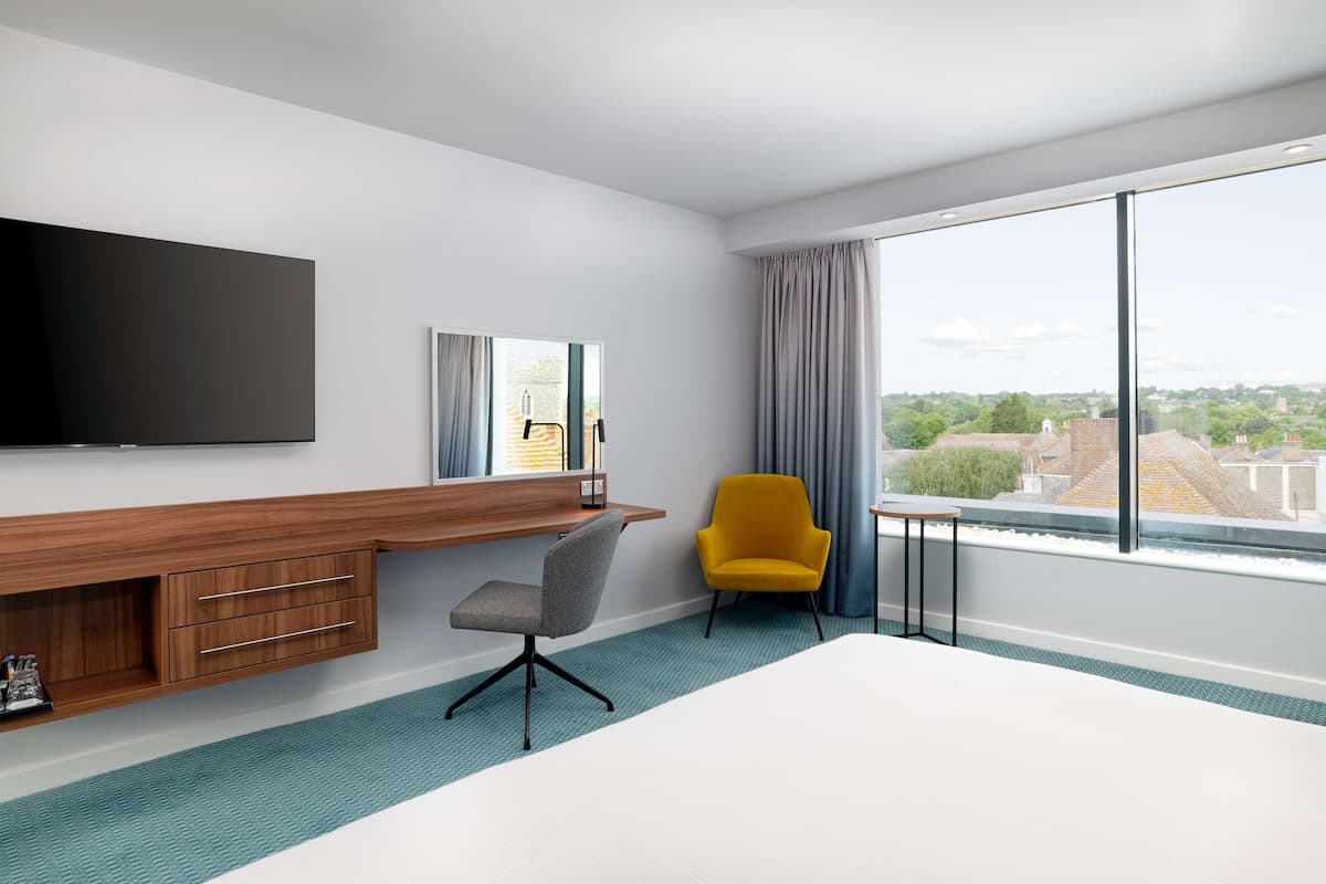 Quarto de hotel com uma cama grande, uma escrivaninha com televisão de tela plana, uma cadeira e uma janela ampla com vista para a cidade.