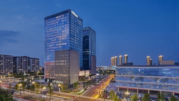 Hotel moderno em arranha-céu com fachada de vidro, cercado por uma área urbana bem iluminada, com outros edifícios e estacionamento.