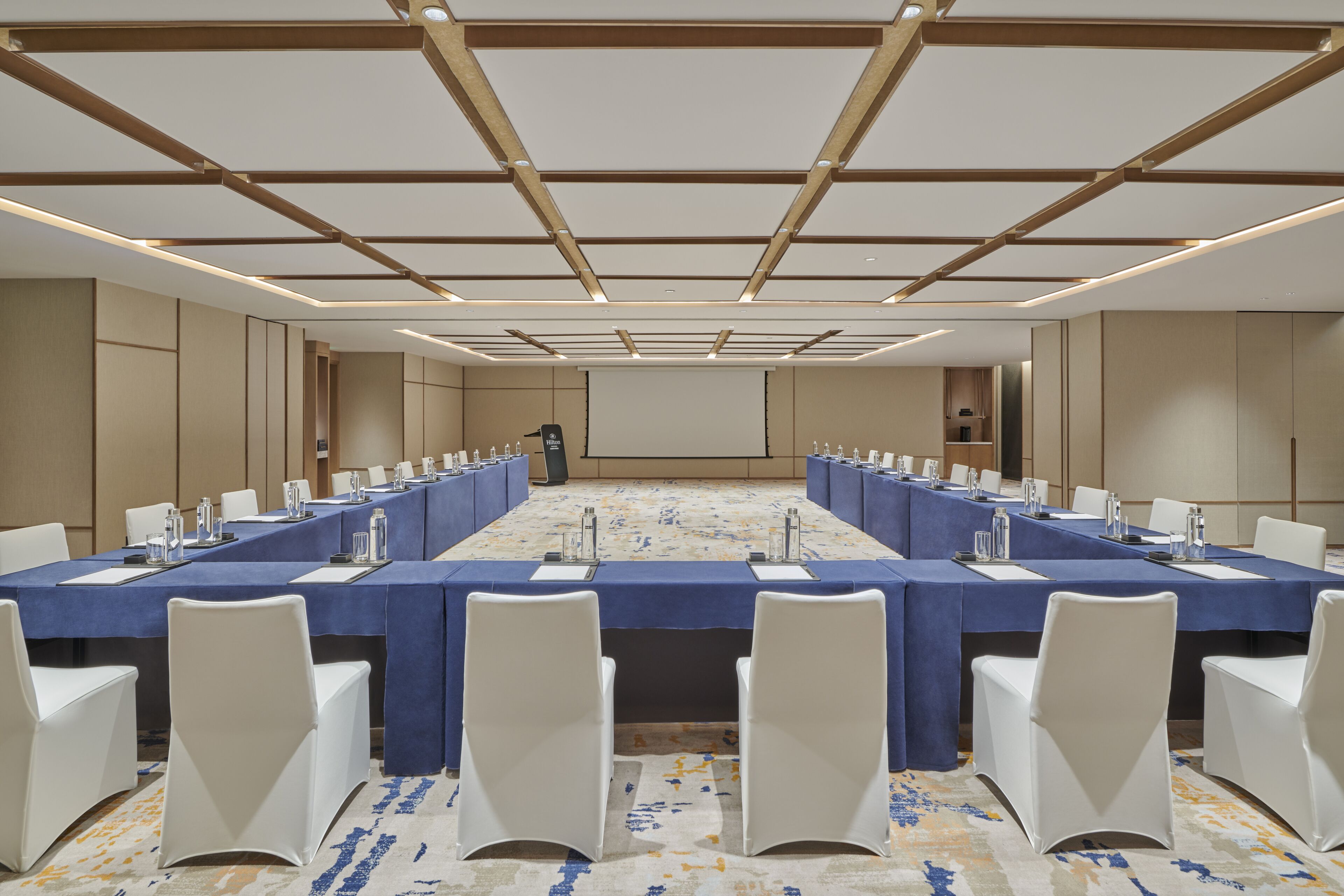 Uma sala de conferências com uma mesa comprida, cadeiras brancas e piso coberto de carpete.