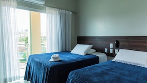 Quarto de hotel com duas camas, uma janela grande com cortinas e vista para vegetação, além de cabeceira de madeira.