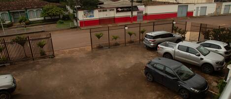 Uma área de estacionamento com vários carros, uma entrada com portão e edifícios ao fundo.