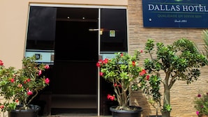 Entrada do Hotel Dallas, com placa, plantas em vasos e varanda de vidro.
