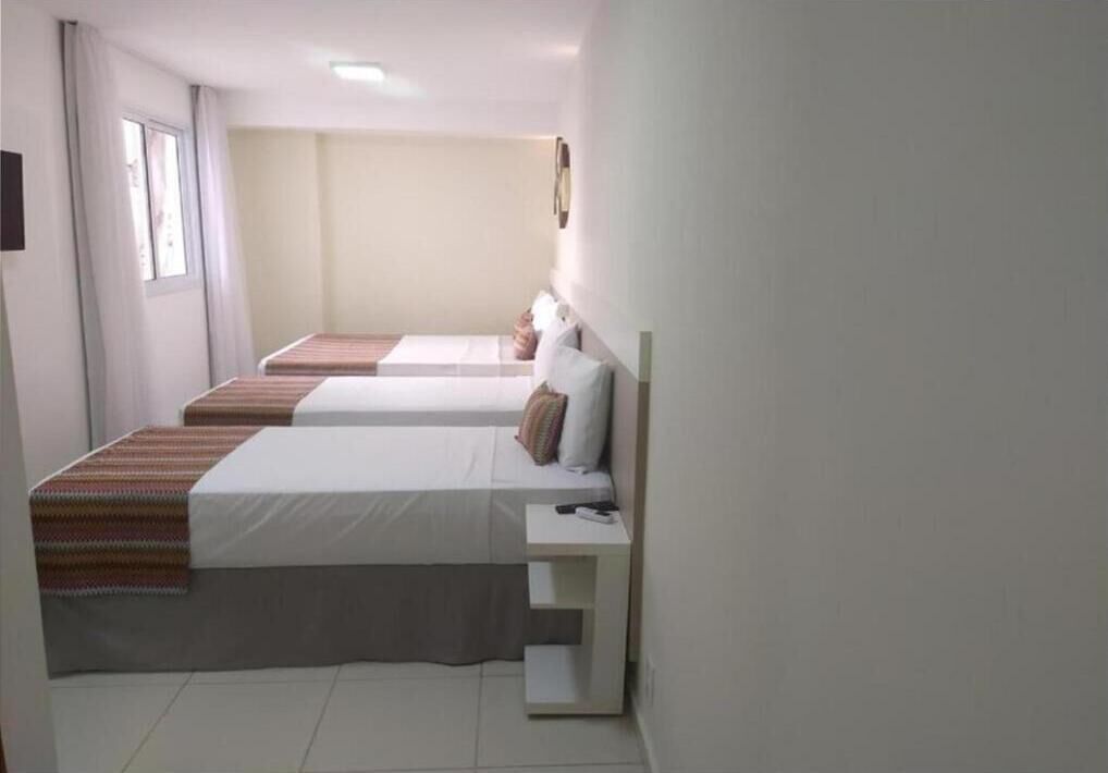 Quarto de hotel com duas camas de solteiro, uma pequena mesa de cabeceira com abajur e uma janela com cortinas.