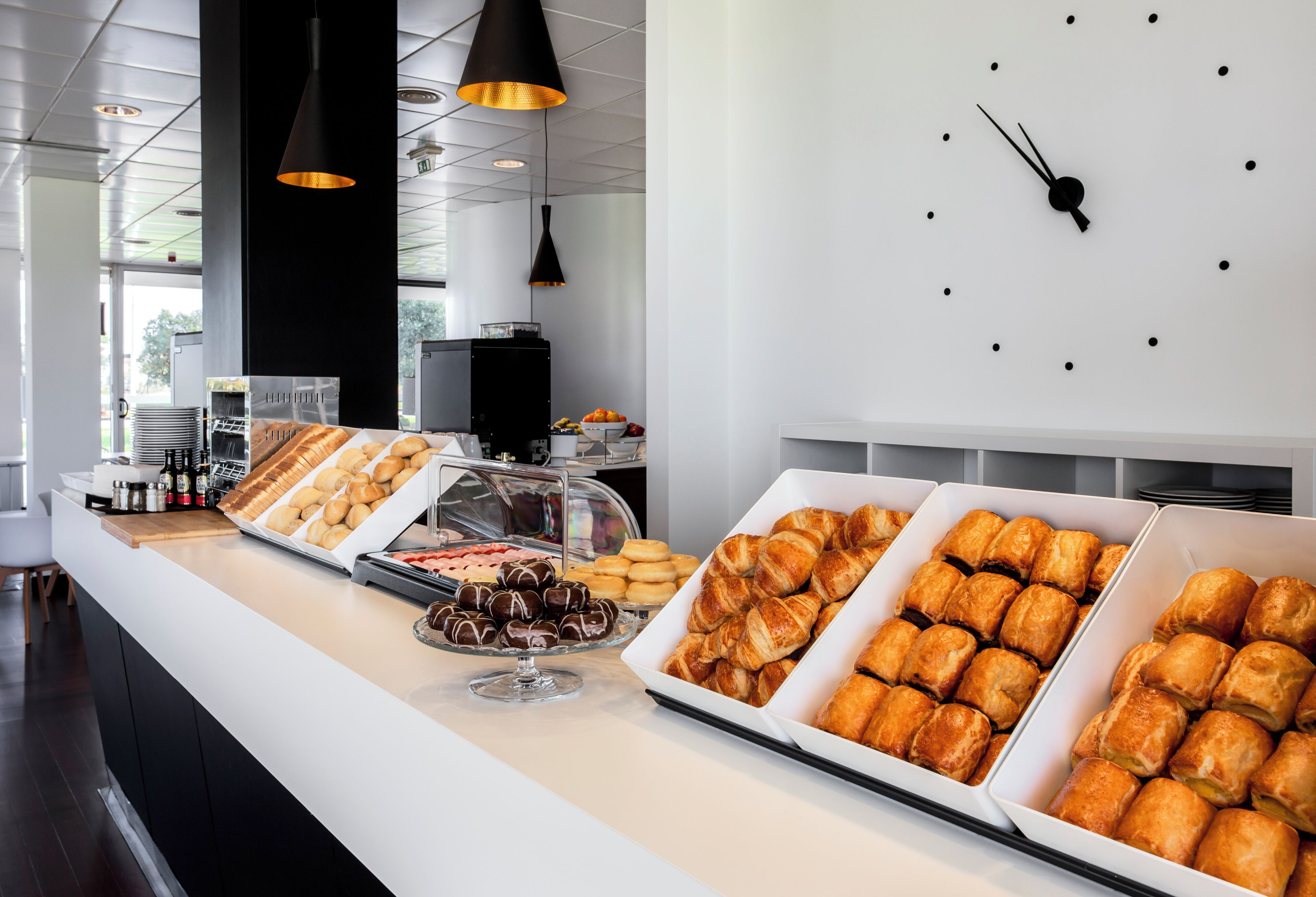 Buffet de café da manhã com doces e produtos de panificação, um relógio na parede e iluminação pendente moderna.