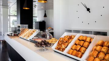 Buffet de café da manhã com doces e produtos de panificação, um relógio na parede e iluminação pendente moderna.